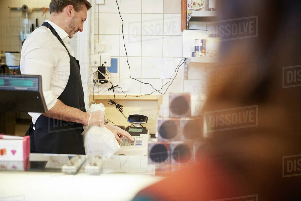 Male cashier holding plastic bag while using cash register in store ...