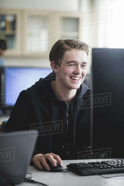 Smiling teenage boy using computer in classroom at high school ...