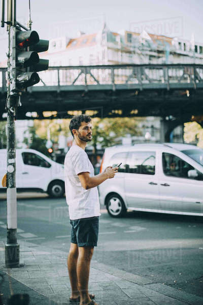 Side view of young man holding mobile phone while standing on sidewalk ...