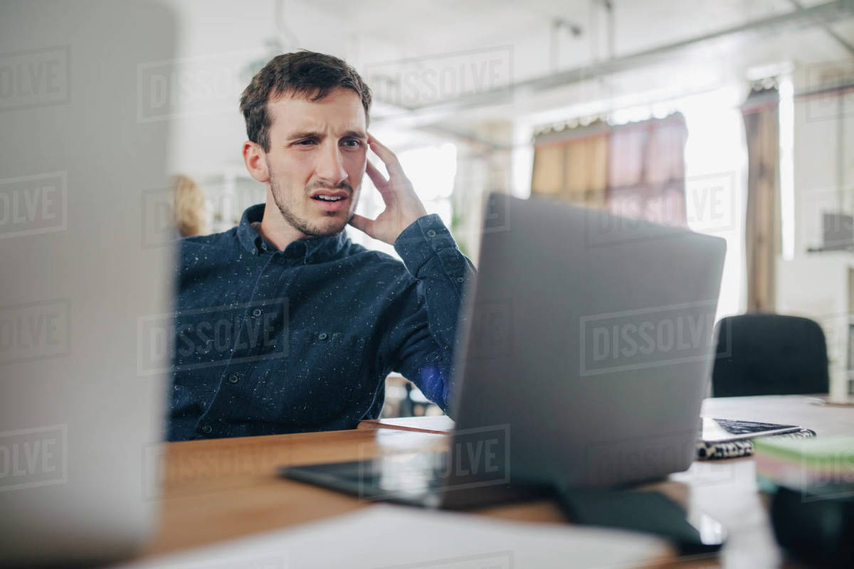 Confused businessman looking at laptop while sitting at desk in office ...