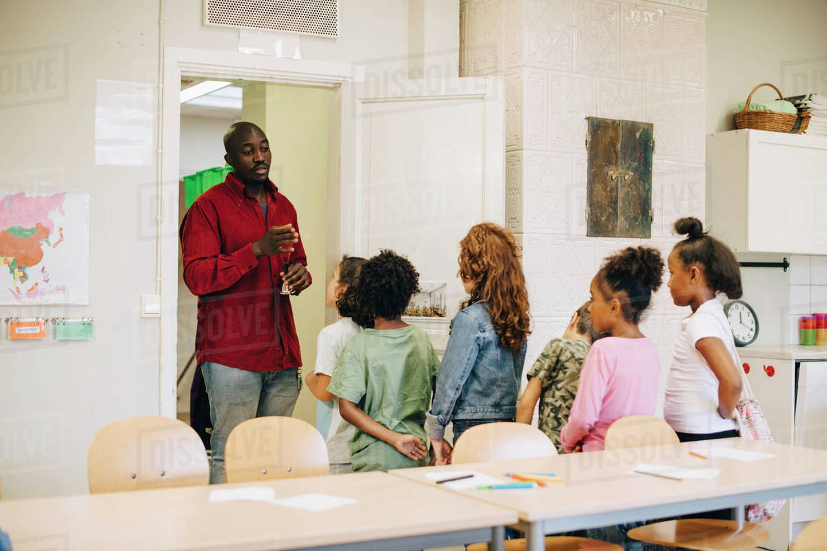 Male teacher guiding and explaining students at doorway in classroom ...