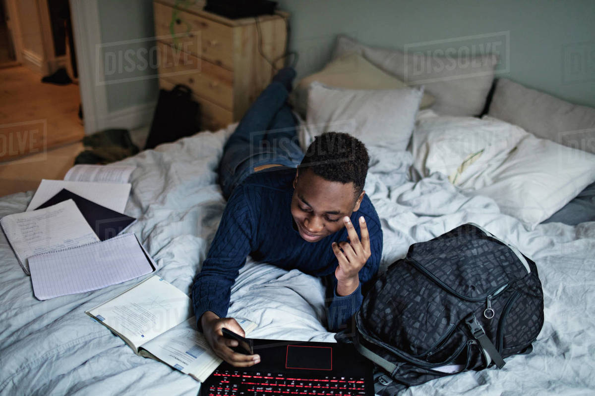 Smiling teenage boy using social media while doing homework on bed at ...