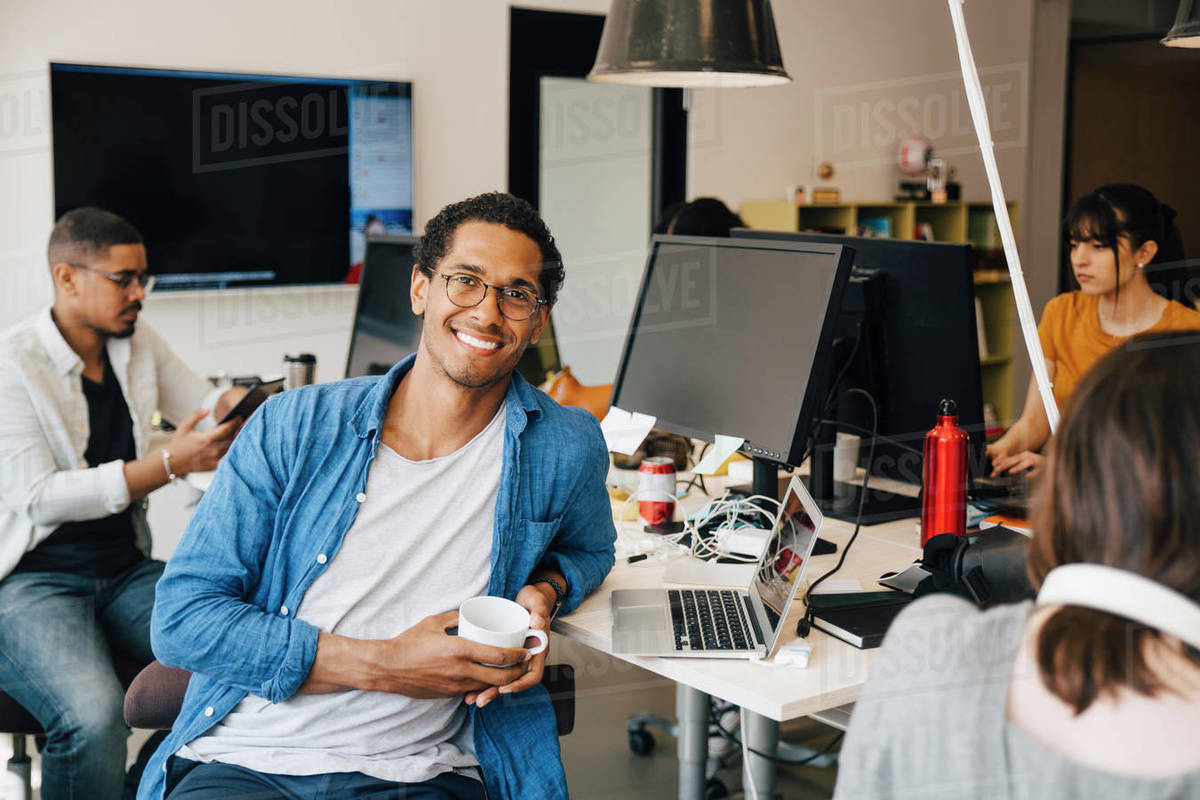 Portrait of male computer programmer sitting by colleagues at desk in ...
