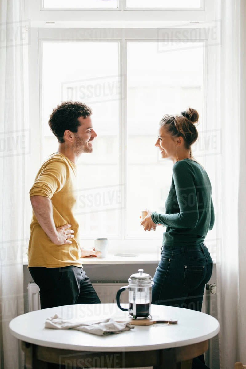 Side view of happy couple talking while standing by window at home ...