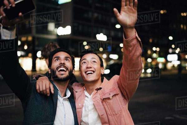 Happy man and male friend looking up while gesturing in city at night ...