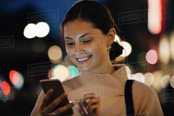 Smiling young woman texting through smart phone in city at night ...