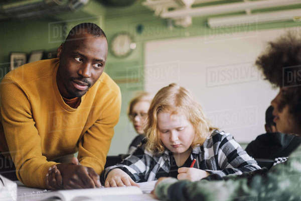 Serious teacher looking at student while leaning at desk in classroom ...