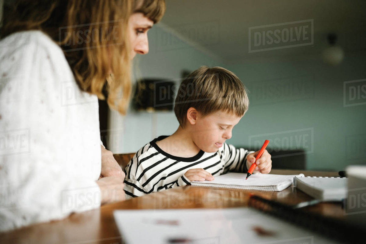 Boy with down syndrome writing in book while sitting by mother at table ...
