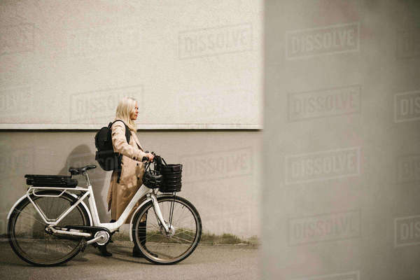 Side view of businesswoman wheeling with electric bicycle by wall ...
