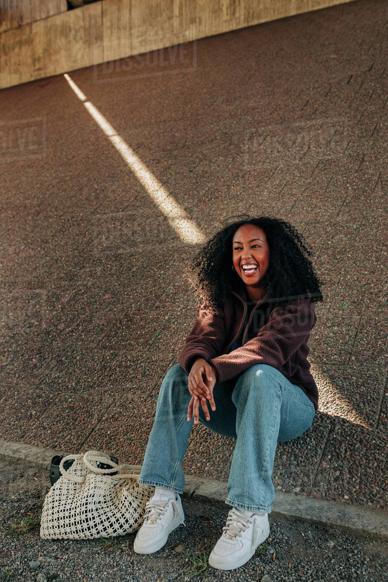 Happy young woman with bag sitting on slope at roadside - Stock Photo ...