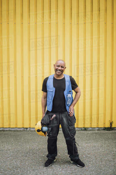 Full length of smiling bald construction worker standing against yellow ...