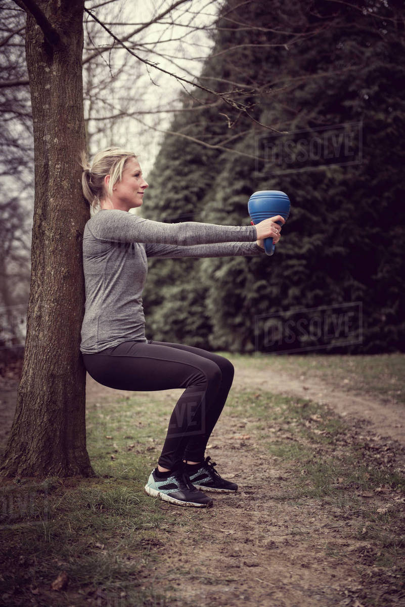 Side view of woman crouching against tree holding kettle bell - Royalty ...