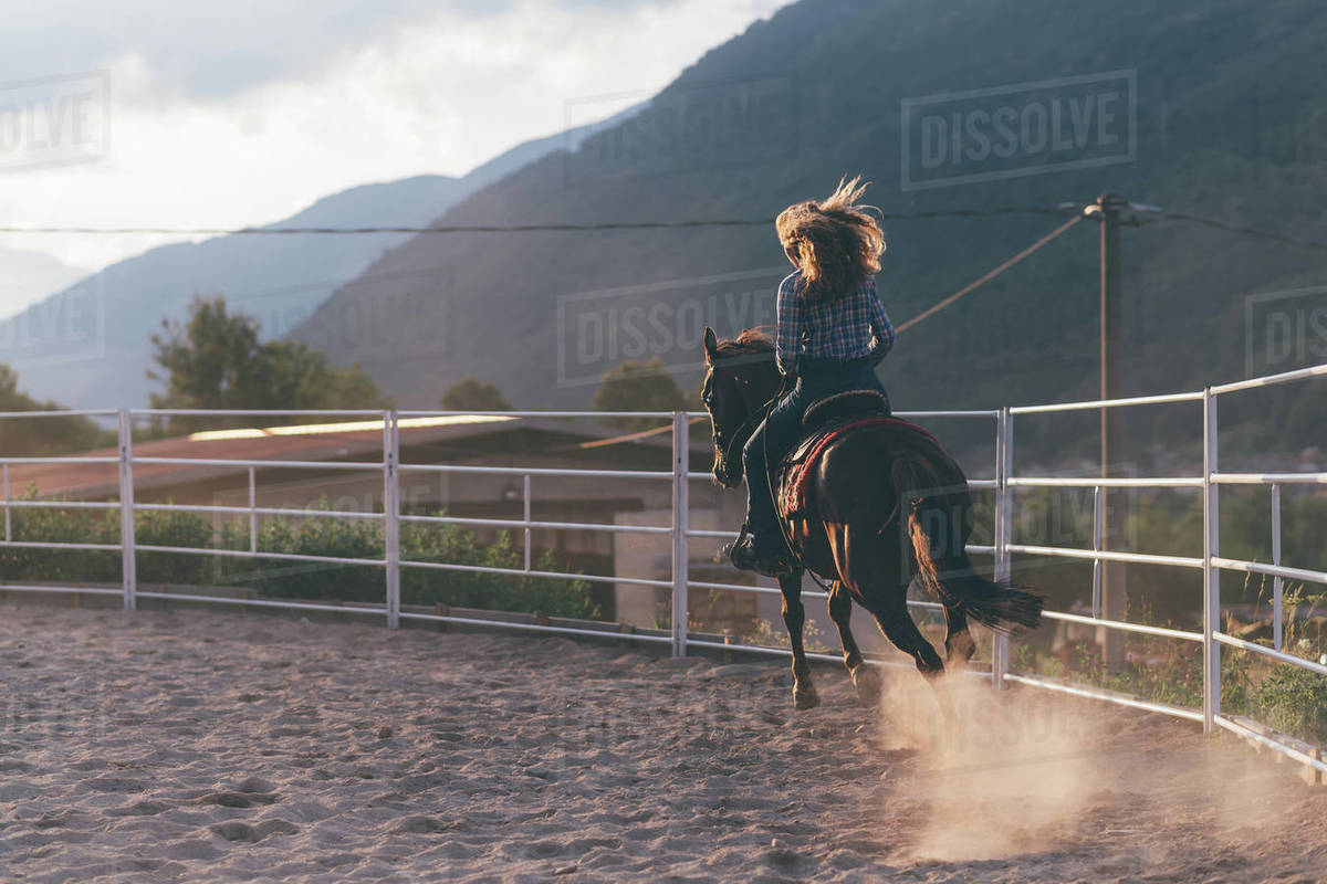 Long haired young woman cantering on horse in rural equestrian arena ...