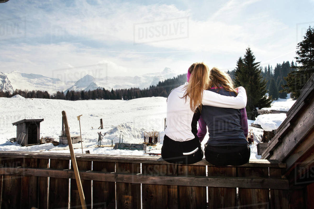 Two teenage girl skiers sitting on cabin roof terrace in snow covered ...