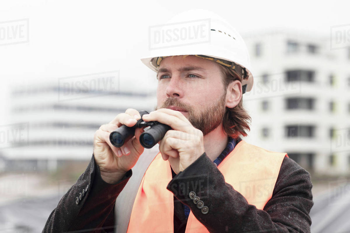 Mid adult male civil engineer holding binoculars on construction site ...