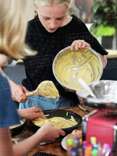 Girl and her sister baking a cake, pouring cake mixture into cake tin ...