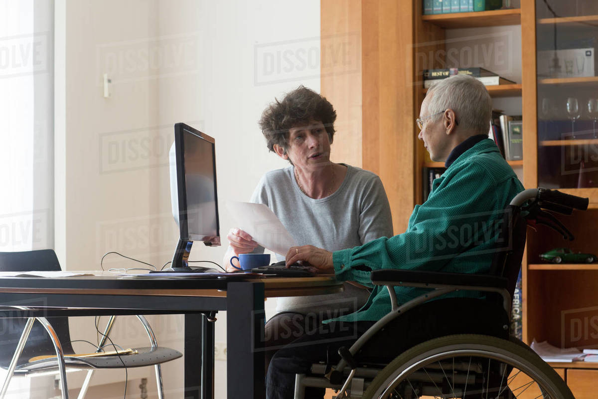 Senior woman sitting in a wheelchair at a computer, helper assisting ...