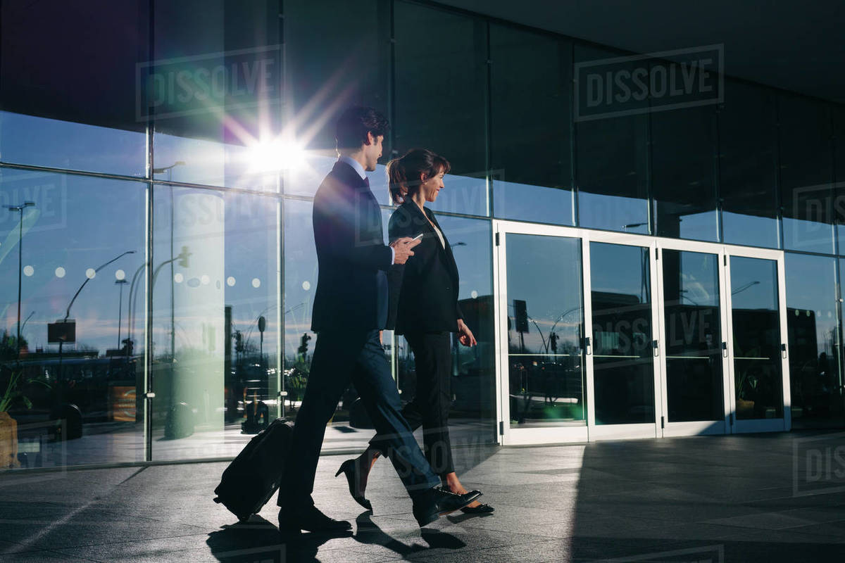 Businessman and businesswoman with wheeled luggage passing glass ...