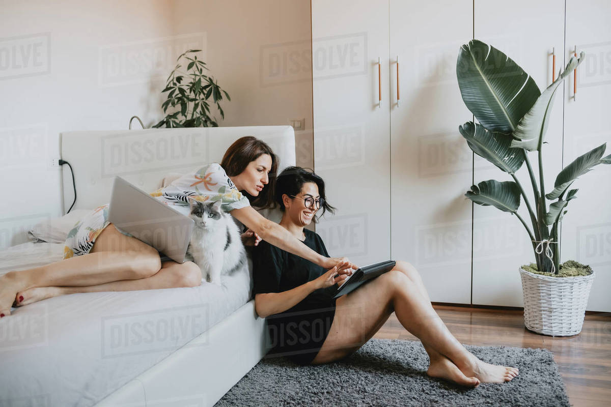 Two women with brown hair sitting on floor and lying on daybed, using ...