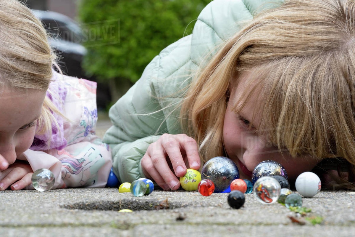 Two children are playing a game with marbles on a paved surface ...