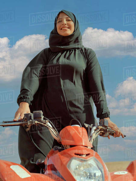 A smiling woman in a blue sweater charges an electric vehicle at sunset ...