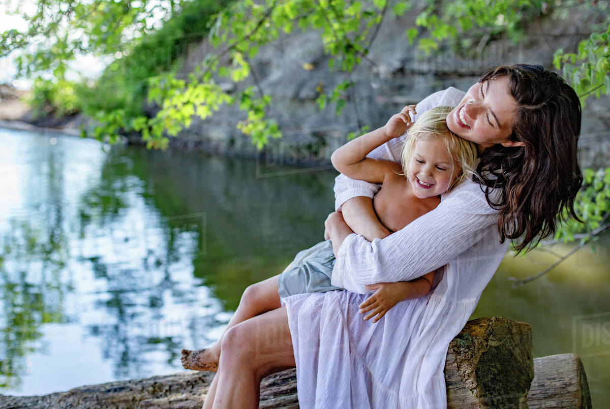 A joyful woman embraces a young boy by a serene lakeside, both smiling and enjoying a sunny day ...