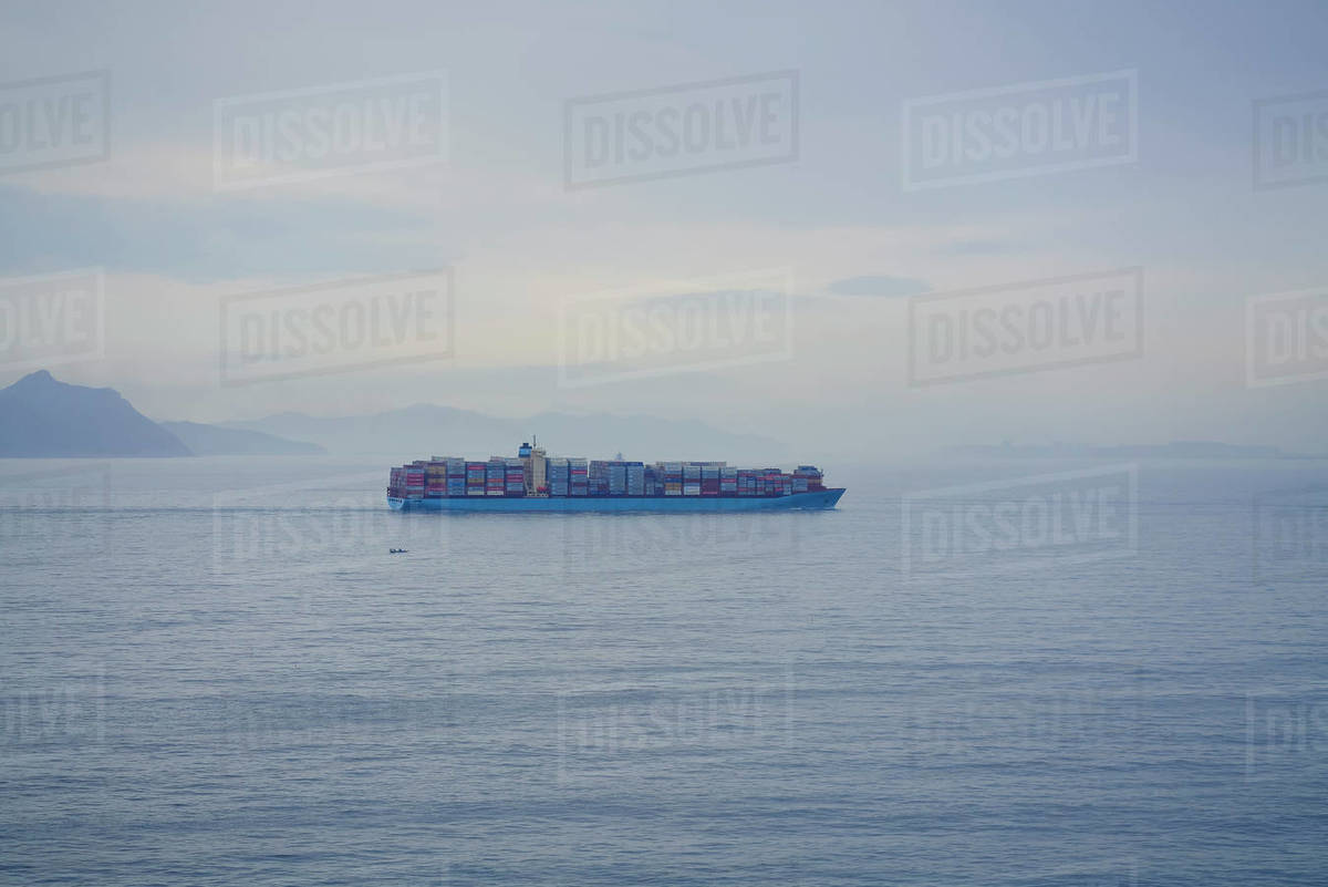 A cargo ship loaded with containers traverses a misty ocean with ...