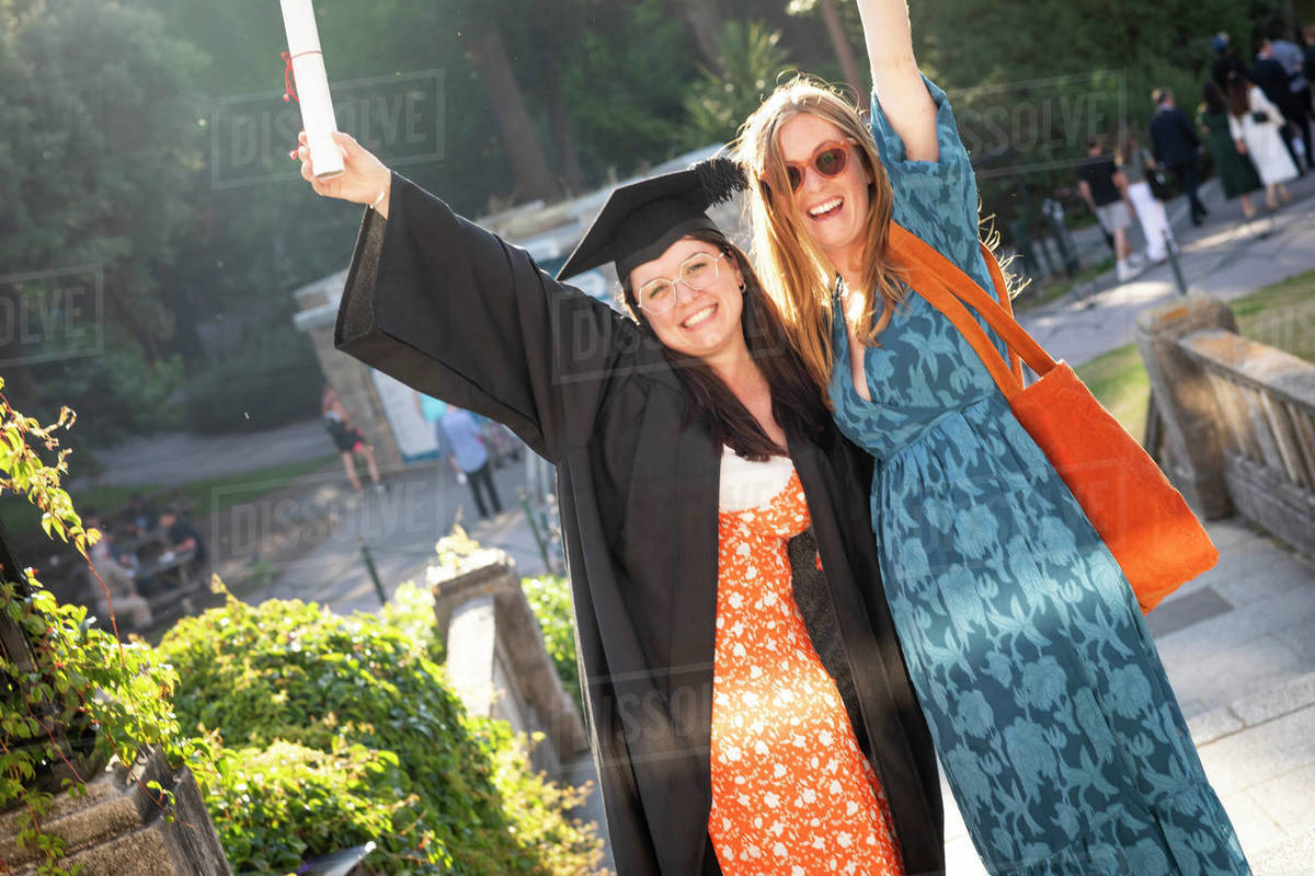 Two women celebrating graduation, one in cap and gown holding a diploma ...