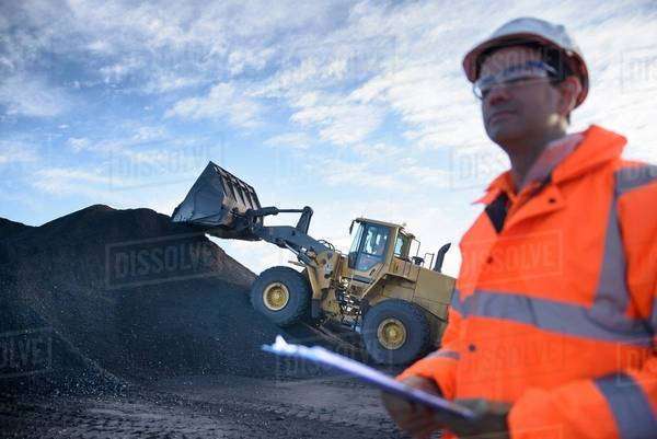 Worker in coal loading station with digger and pile of coal at surface ...