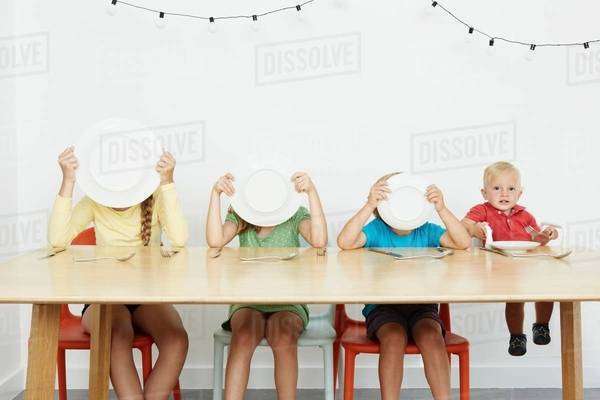 Four children sitting at table, three covering faces with plates ...