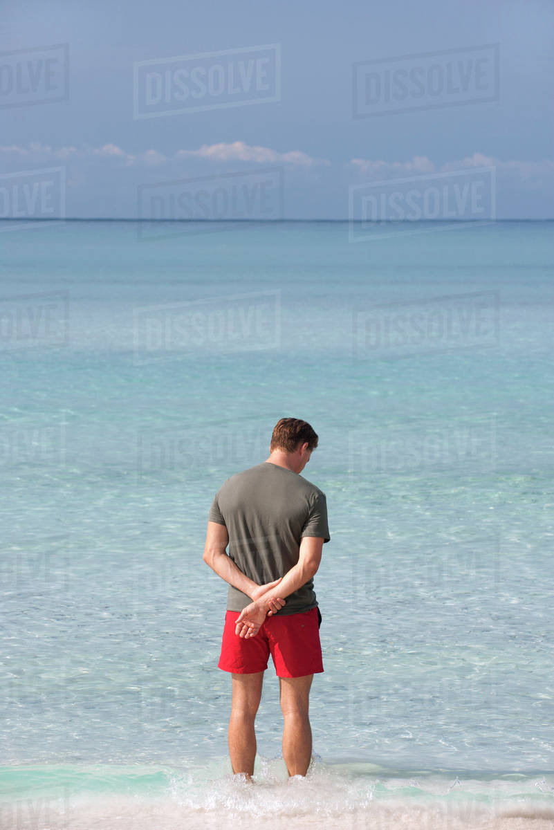 Man standing ankle deep in water contemplating ocean, rear view ...