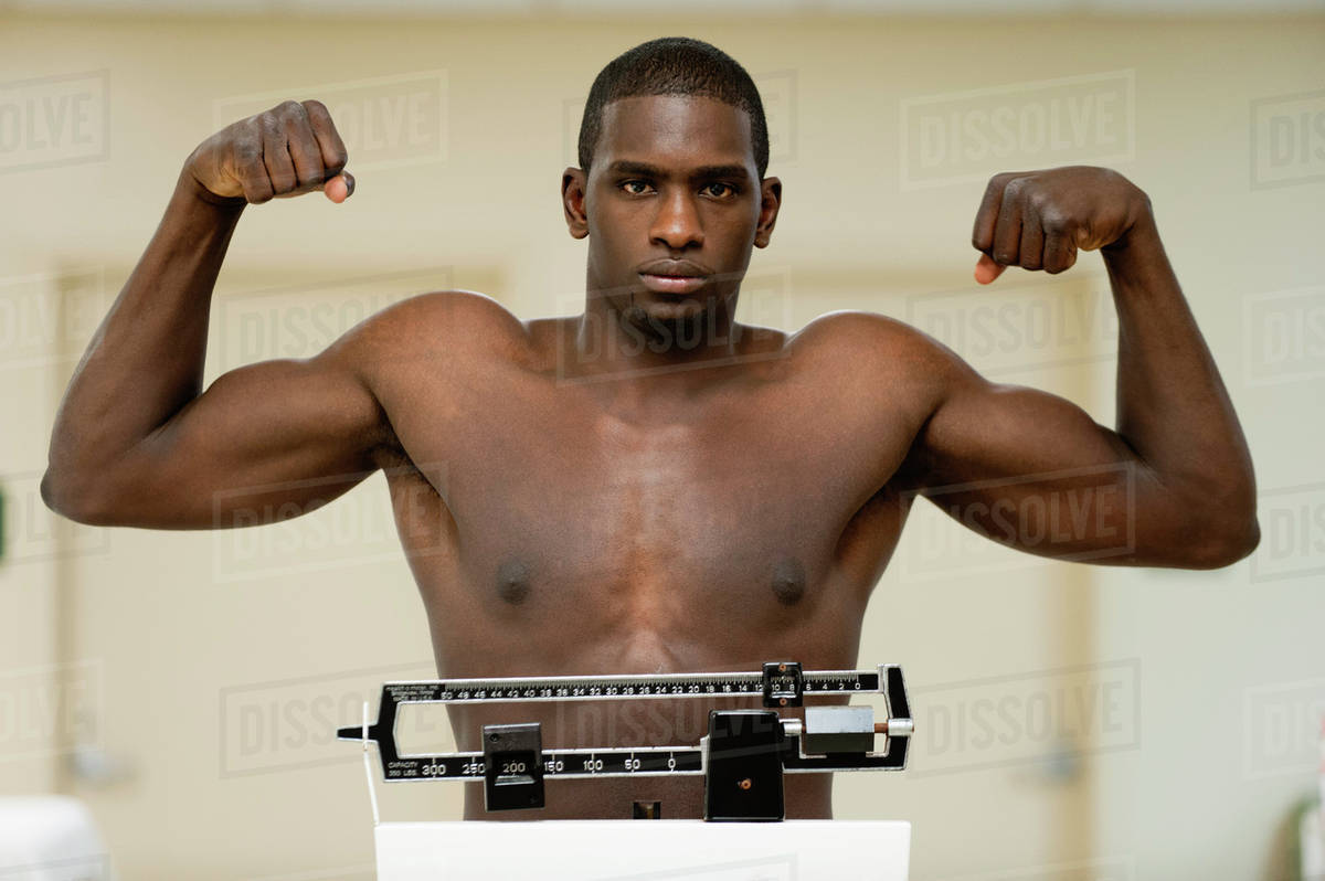 Young man flexing muscle on weight scale, portrait - Stock Photo - Dissolve