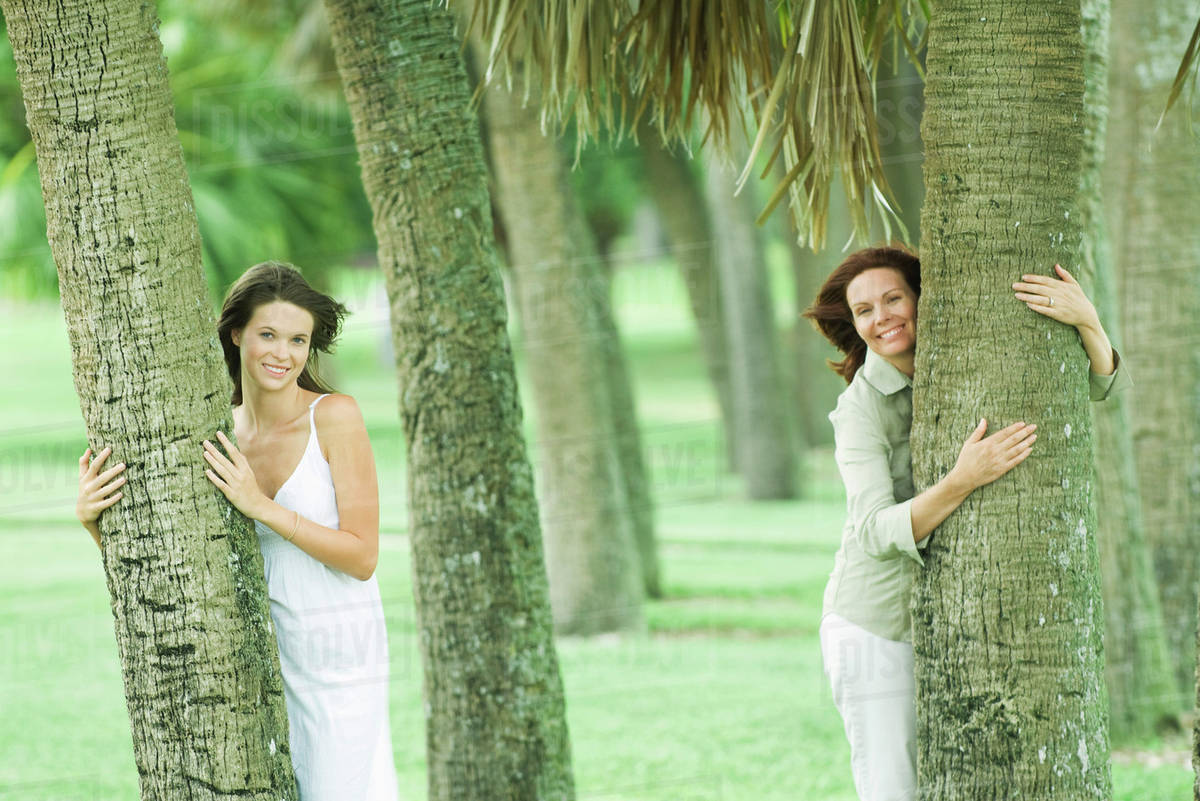 Mother and teen daughter hugging tree trunks, smiling at camera ...