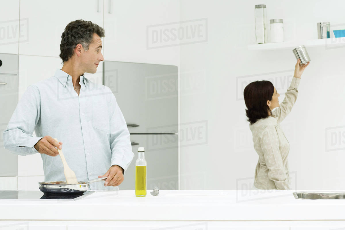 Couple in kitchen, man cooking at stove, looking over shoulder, wife ...