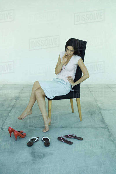 Young woman sitting in chair, looking down at three pairs of shoes ...
