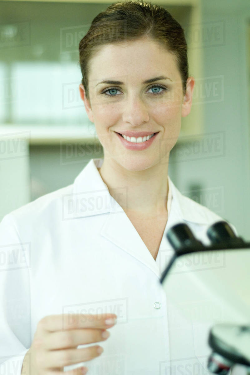 Female lab worker with microscope, holding up slide, smiling at camera ...