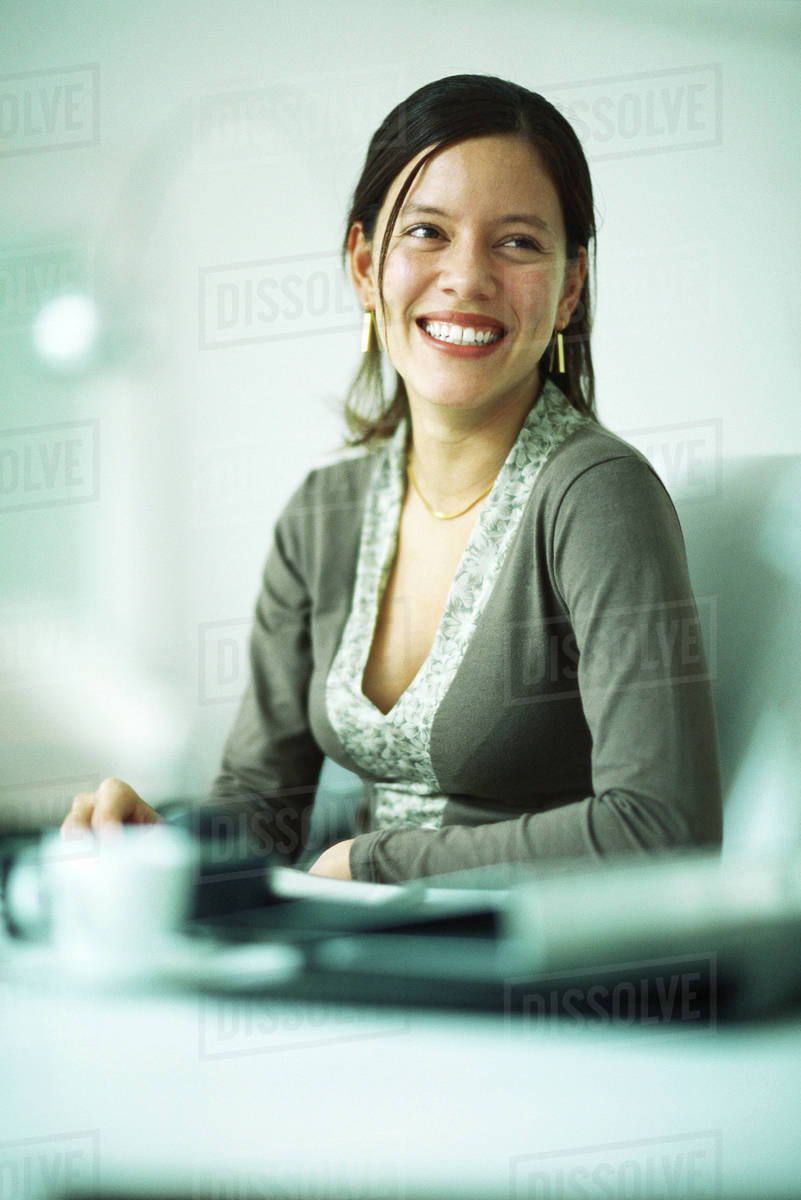 Woman sitting at desk, smiling, looking away - Stock Photo - Dissolve