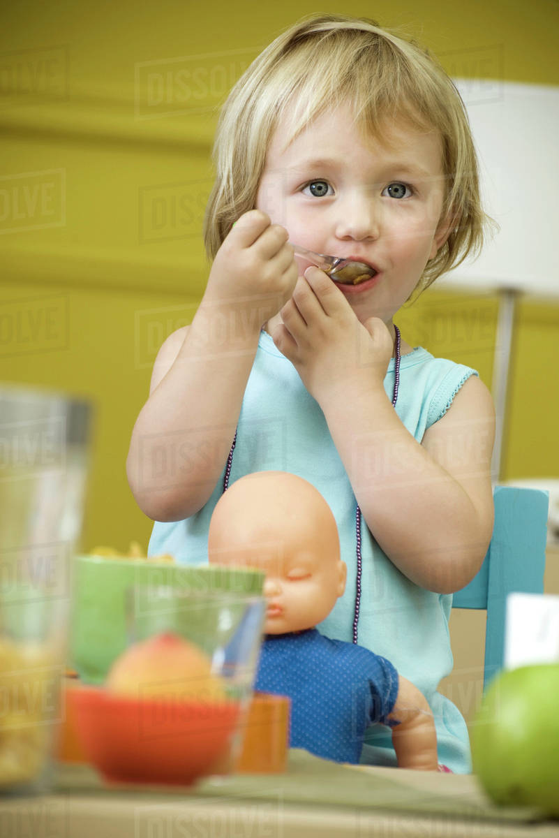 Toddler girl eating at table, baby doll in lap - Royalty-free Stock ...
