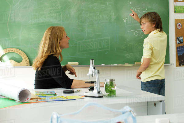 Boy solving math problem on blackboard, looking at teacher for ...