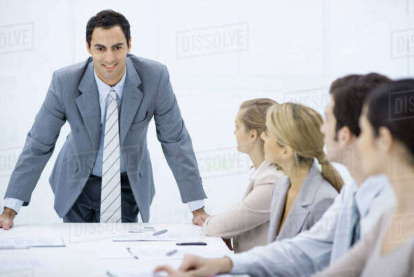 Businessman leaning over table addressing colleagues, smiling at camera ...