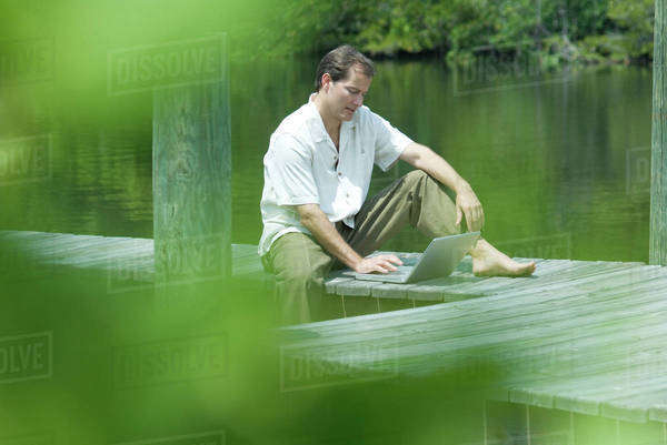 Man sitting on dock using laptop computer, view through foliage ...