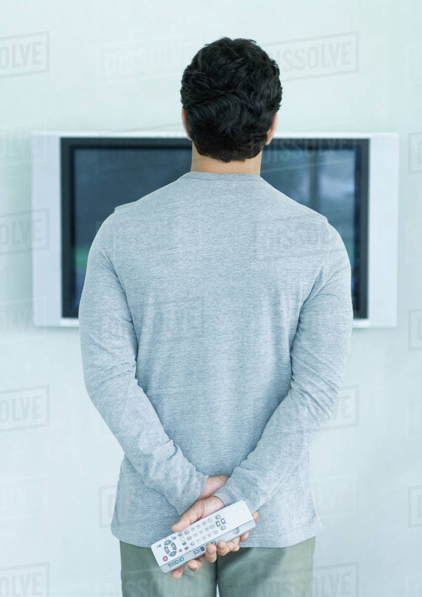Man standing in front of widescreen TV, holding two remote controls ...