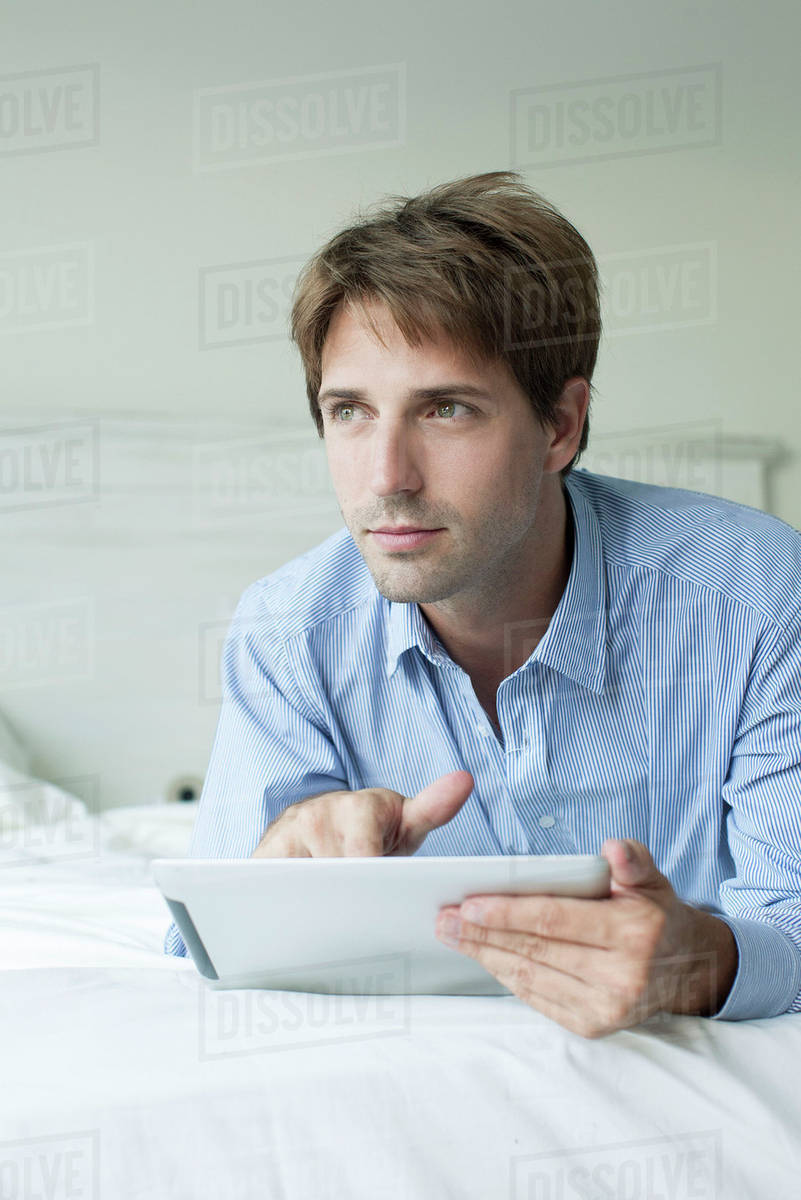 Man lying in bed using digital tablet, looking away with contemplative ...