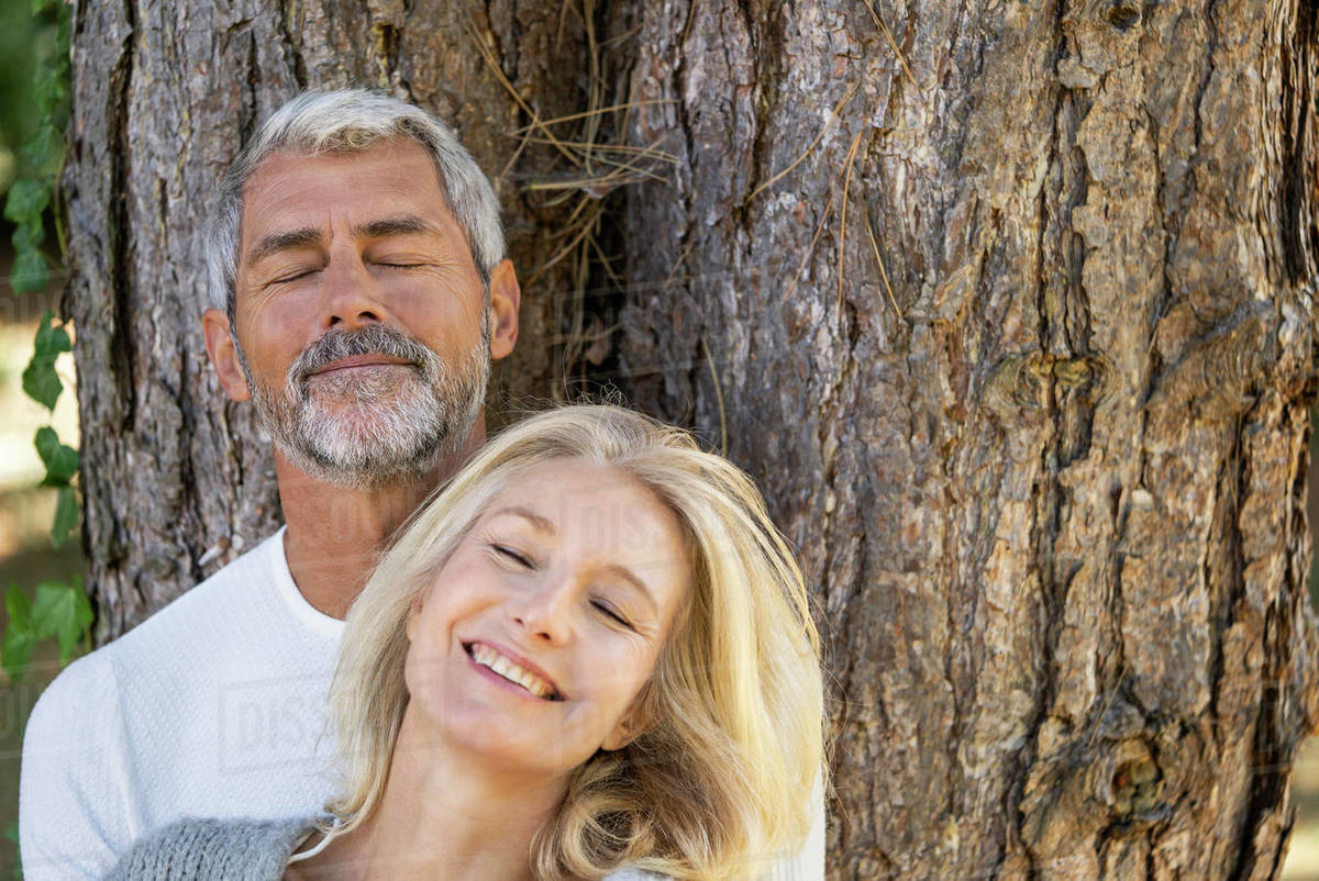 Smiling mature couple with eyes closed leaning on tree trunk - Stock ...