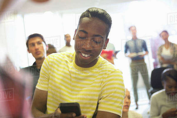 University student texting in classroom, people standing in background ...