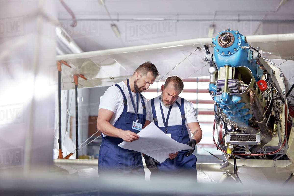 Male engineer mechanics examining plans, fixing airplane in hangar ...