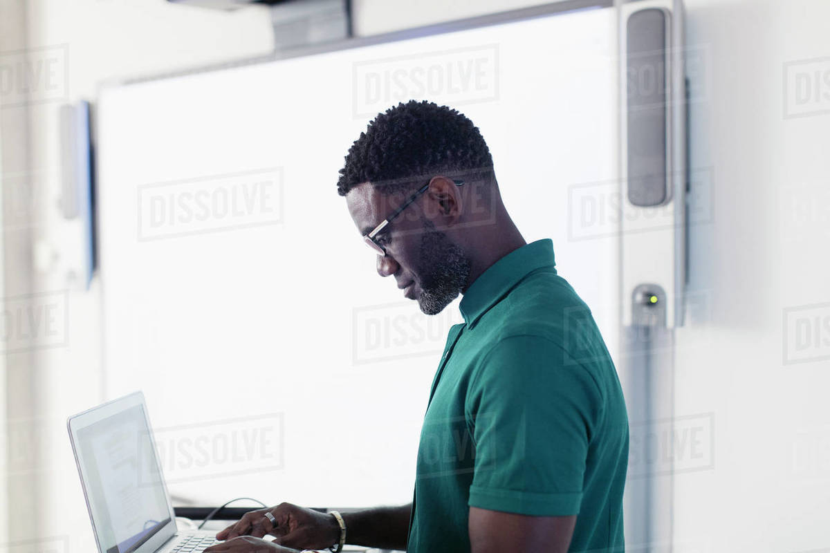 Male professor using laptop next to projection screen in classroom ...