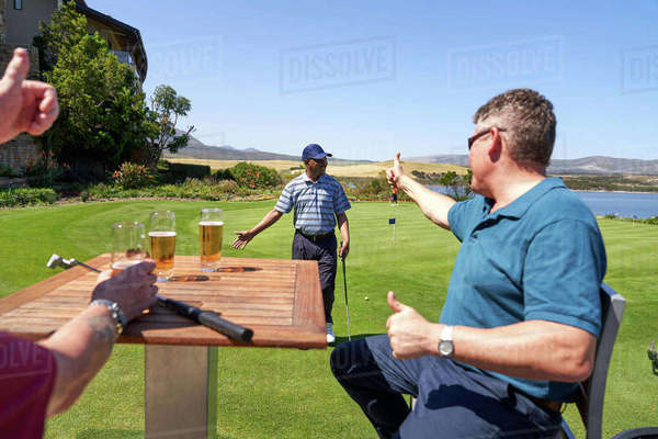Male golfers drinking beer cheering friend on practice putting green ...