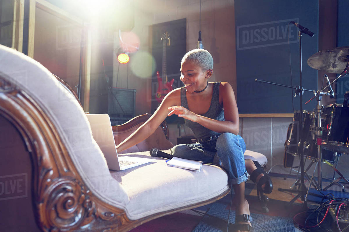 Smiling female musician song writing at laptop in recording studio ...