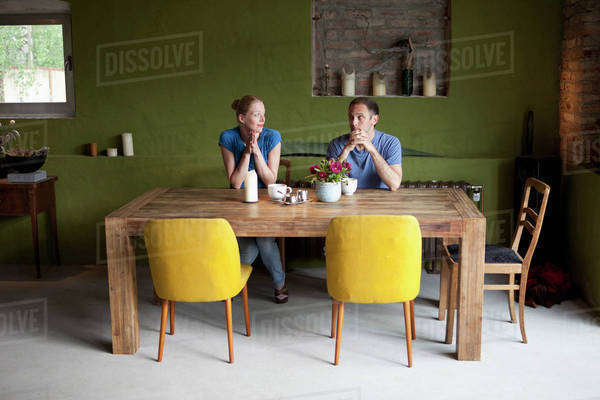 A couple sitting at a dining table looking at each other uncertainly ...